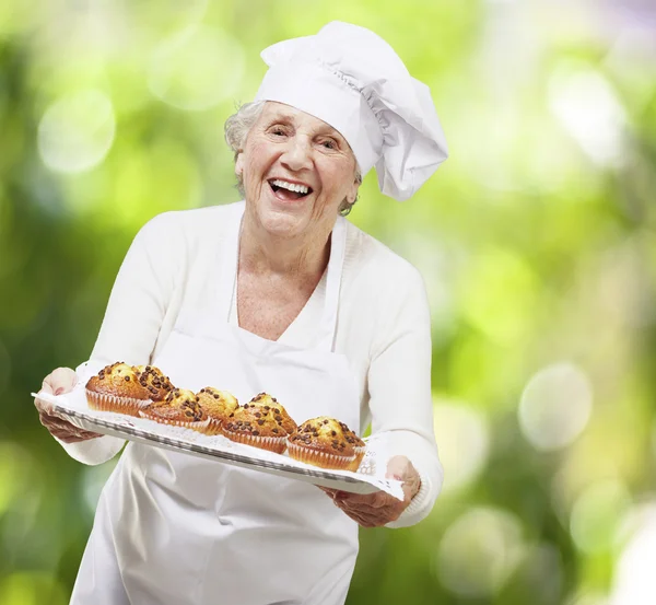 Senior woman cook holding a tray with muffins against a nature b ...